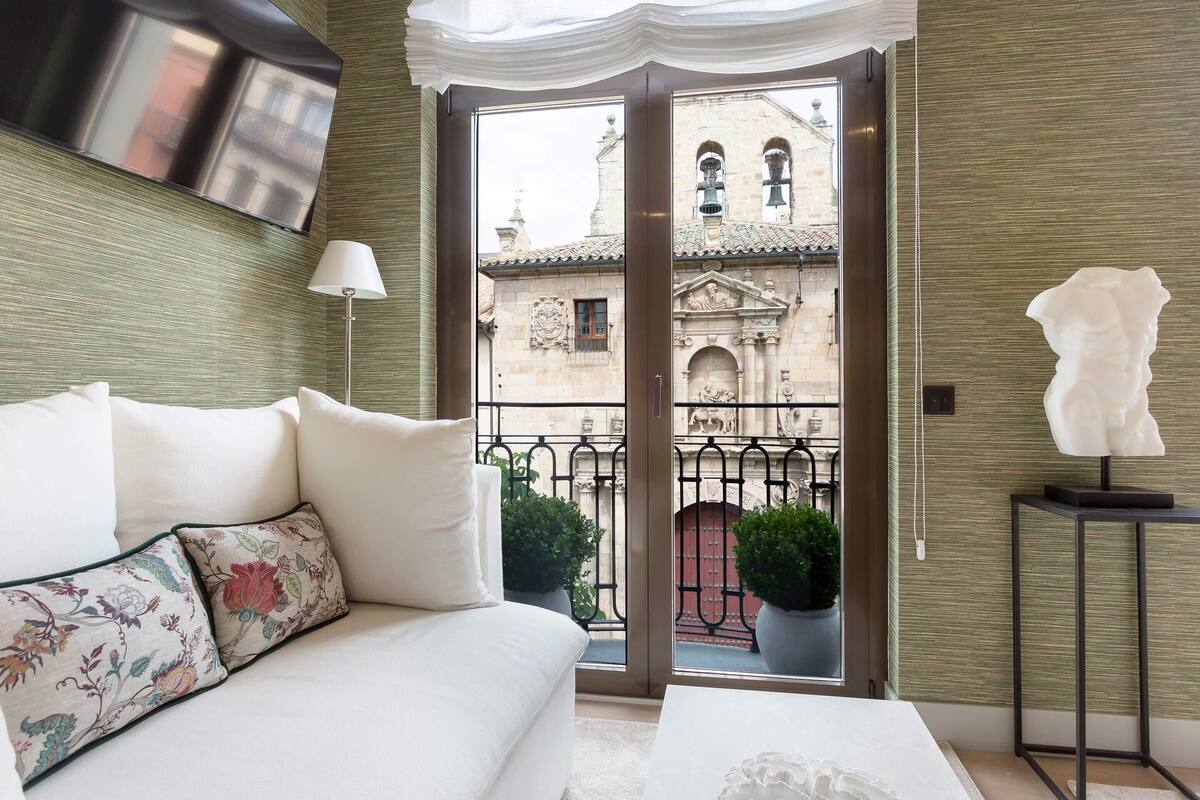 Quarto de hotel moderno com sofá, televisão de tela plana, varanda com plantas em vasos e vista para edifícios históricos.