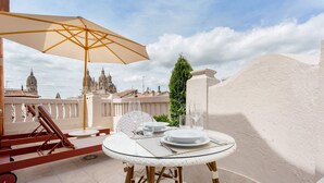 Uma mesa de jantar com tampo de vidro, rodeada por cadeiras, sob um grande guarda-sol, com vista para a cidade ao fundo.