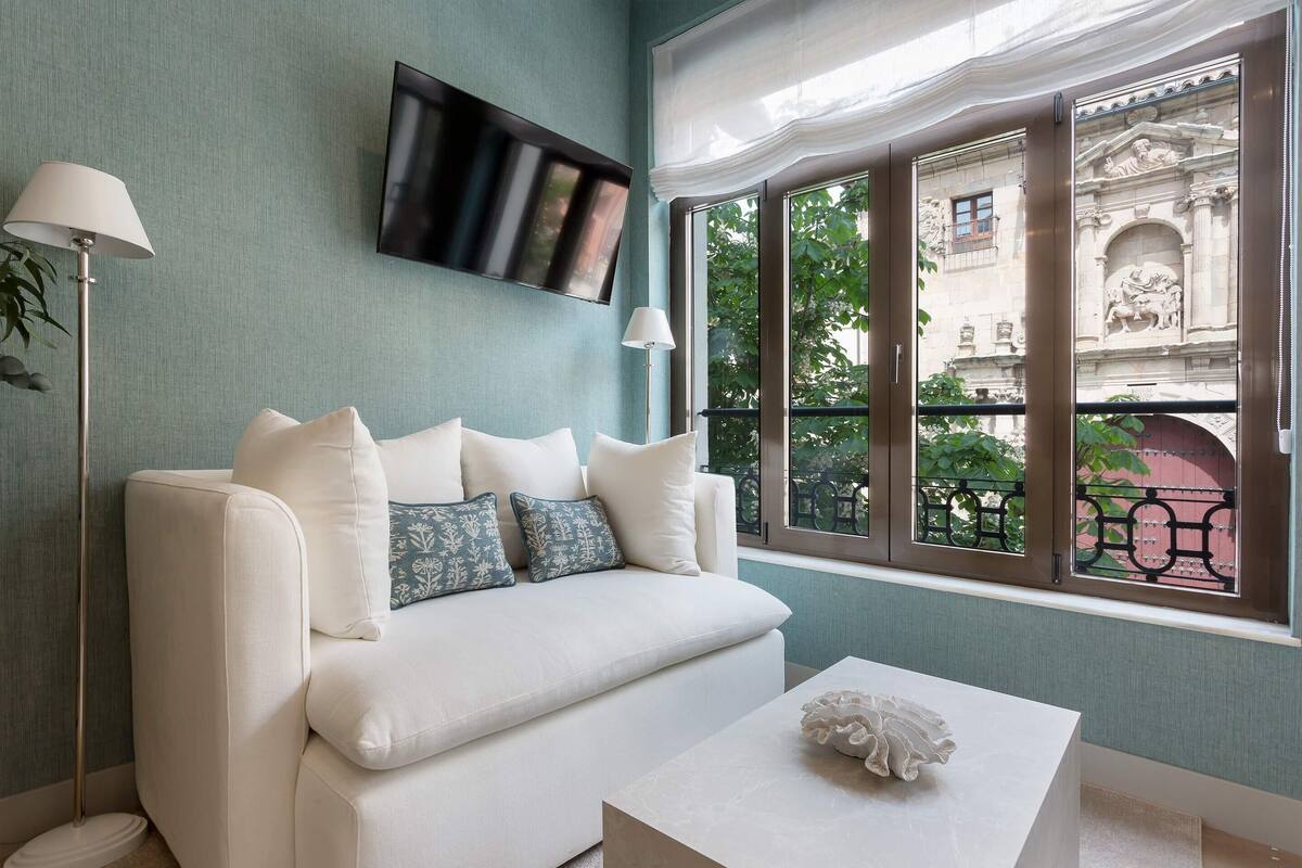Sala de estar moderna com um sofá branco, uma mesa de centro e uma grande janela com vista para um edifício.