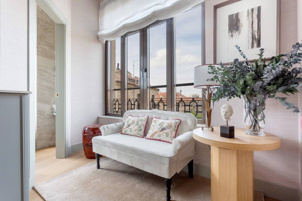 Quarto com sofá branco, mesa redonda de madeira, um vaso com flores e vista para edifícios através de janelas amplas.