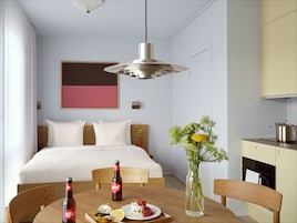 Quarto de hotel com cama, mesa de jantar com uma garrafa e um prato de comida, vaso com flores e cozinha.