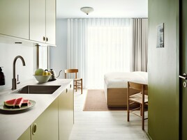 Quarto de hotel compacto com cozinha compacta, uma cama, uma cadeira e uma janela com cortinas.