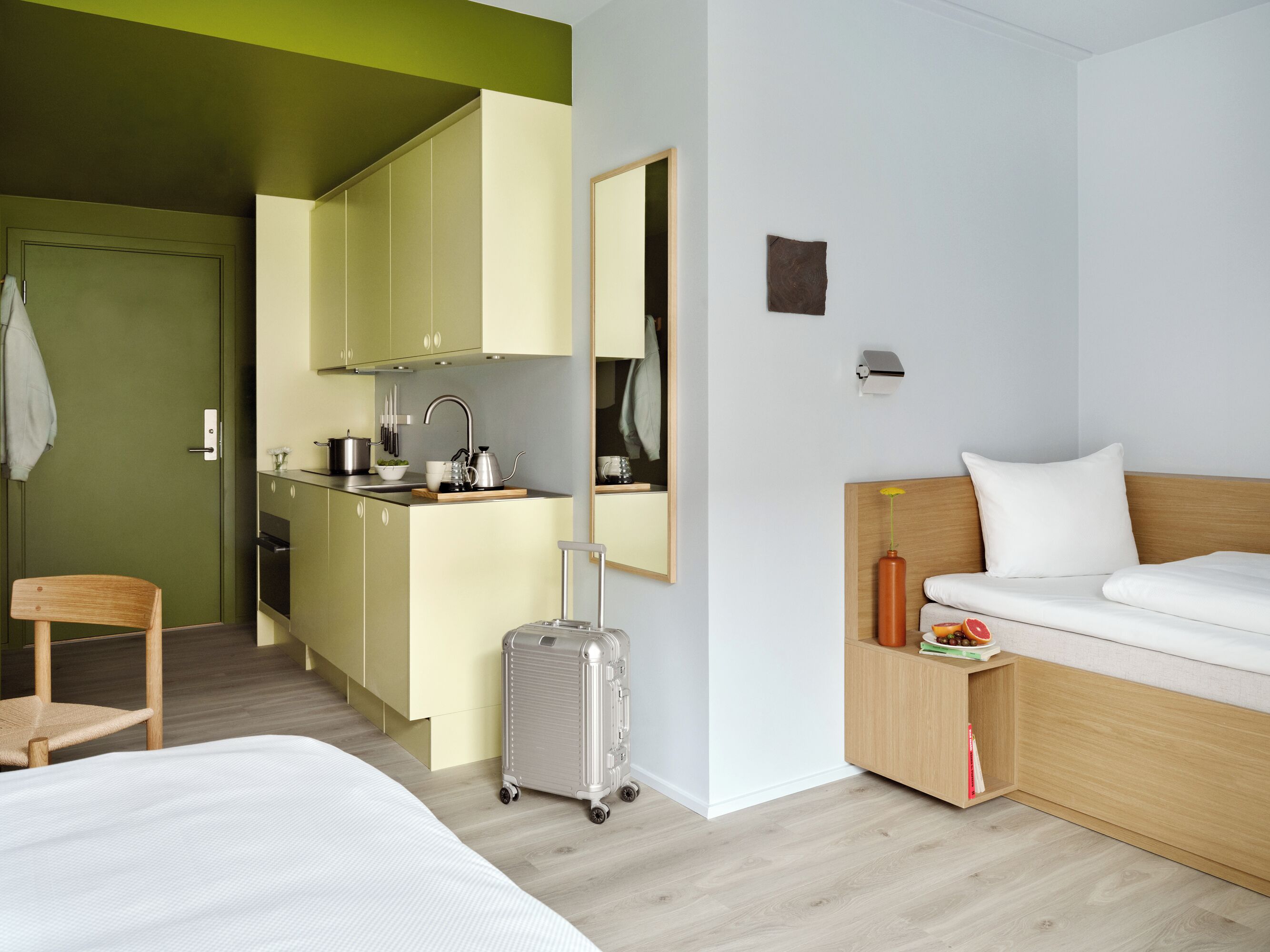 Quarto de hotel compacto com cozinha compacta, uma cama, uma cadeira e uma mala.