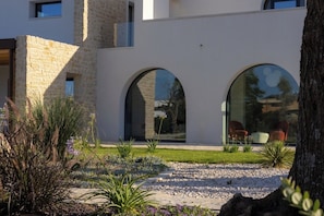 Um edifício moderno com fachada de pedra, grandes janelas em arco e um jardim ajardinado com diversas plantas.