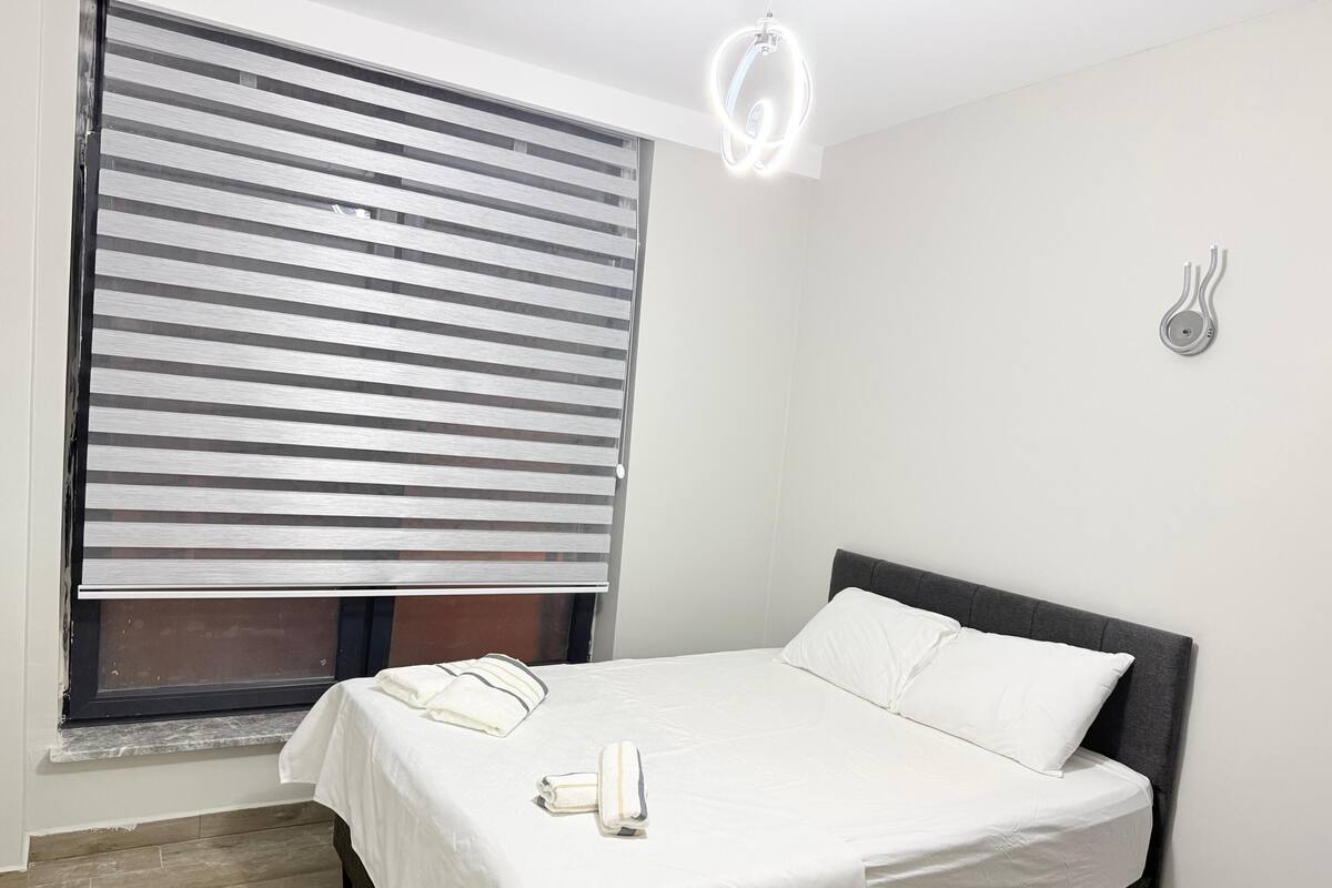 Quarto com cama, janela com persianas, toalha e luminária fixada na parede.