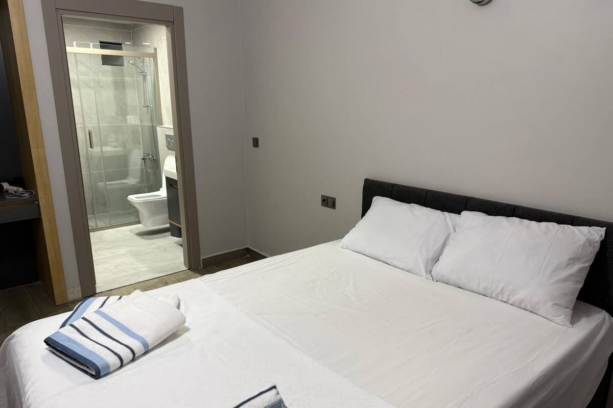 Quarto de hotel com uma cama, banheiro visível por uma porta aberta e um interruptor de luz fixado na parede.