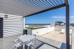 Terraço no telhado com mesa e cadeiras, vista para o mar e área coberta com pérgola.