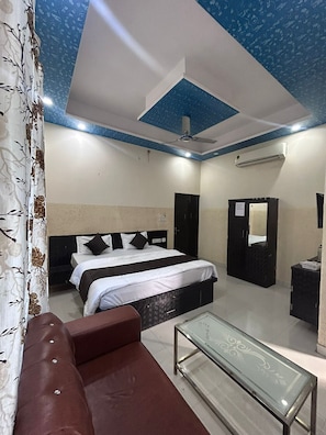 Quarto de hotel com uma cama, um sofá vermelho, uma mesa de centro de vidro, um aparelho de ar condicionado e um ventilador de teto.