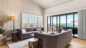 Uma sala de estar moderna com um sofá cinzento, uma mesa de centro e uma luminária. Há grandes janelas com persianas, vista para o mar e uma varanda com mobiliário exterior.