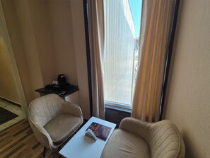 Quarto de hotel com área de estar, uma mesinha com uma revista, cafeteira e janela com cortinas.