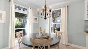 Uma sala de jantar com uma mesa redonda, cadeiras, um candelabro e janelas amplas com cortinas.