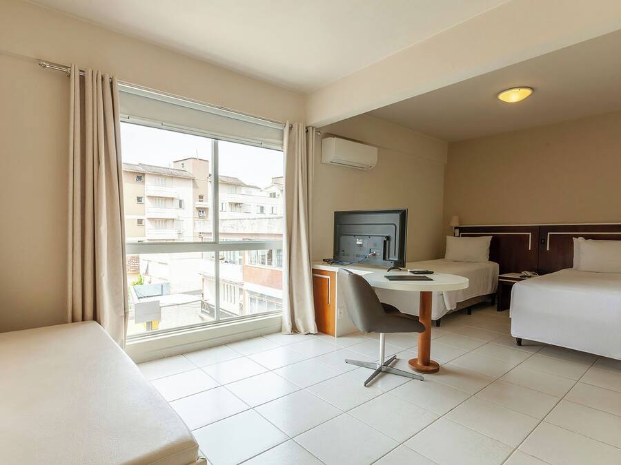 Quarto de hotel com cama, escrivaninha com cadeira, televisão e janela com cortinas.