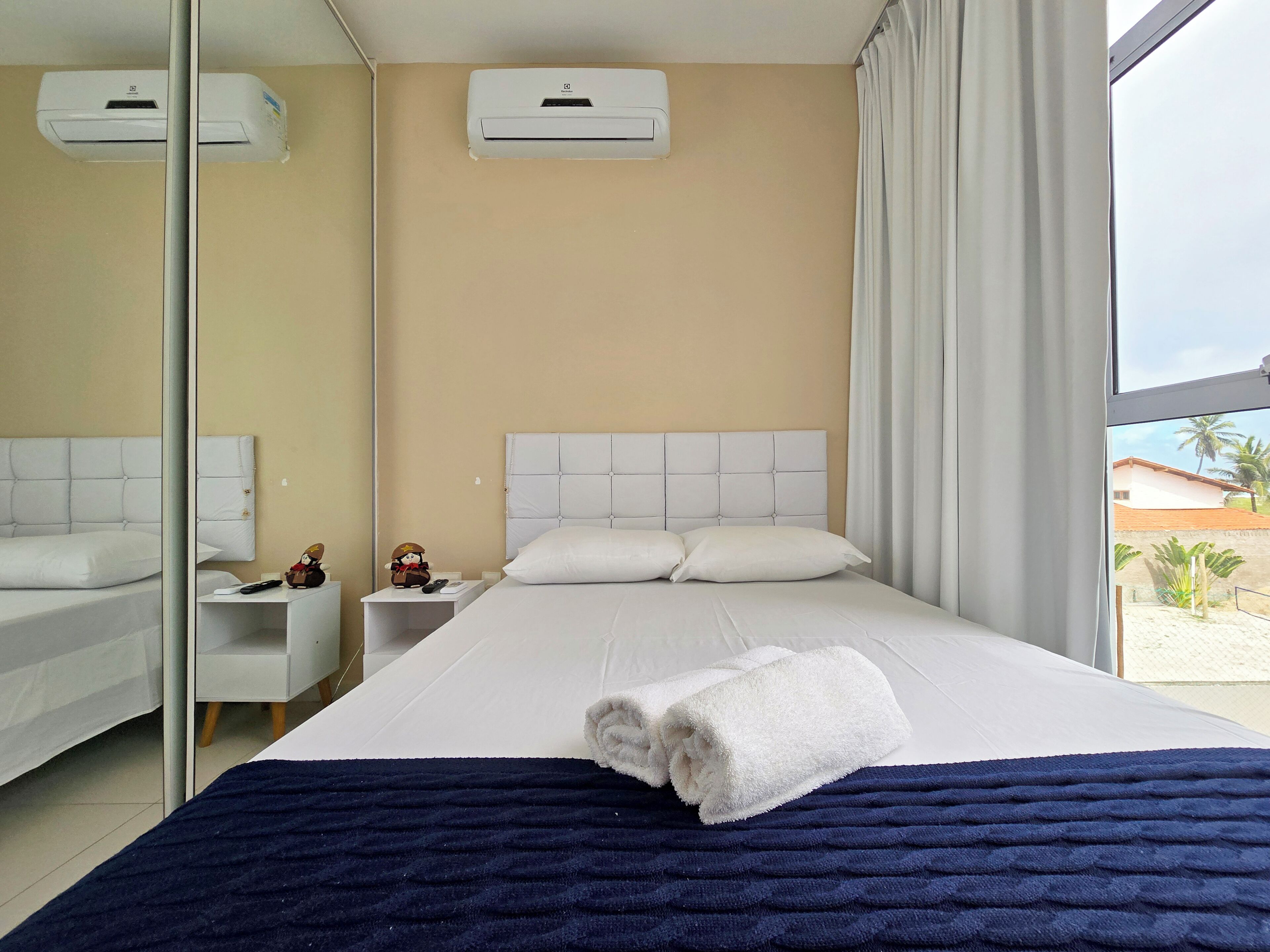 Quarto de hotel com cama, mesa de cabeceira, espelho, aparelho de ar condicionado e vista para palmeiras.