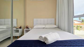 Quarto de hotel com cama, mesa de cabeceira, espelho, aparelho de ar condicionado e vista para palmeiras.