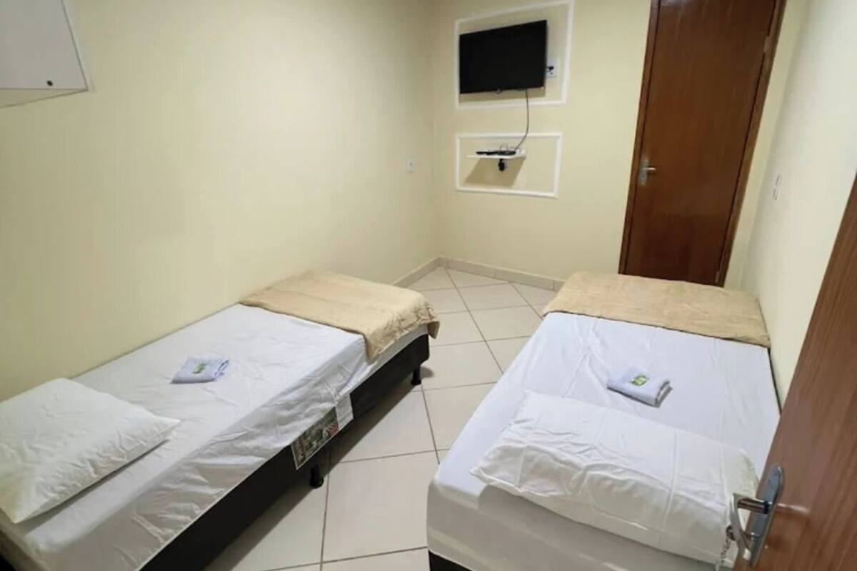 Quarto pequeno e limpo com duas camas de solteiro, um ventilador de teto, uma TV fixada na parede e uma porta de madeira.