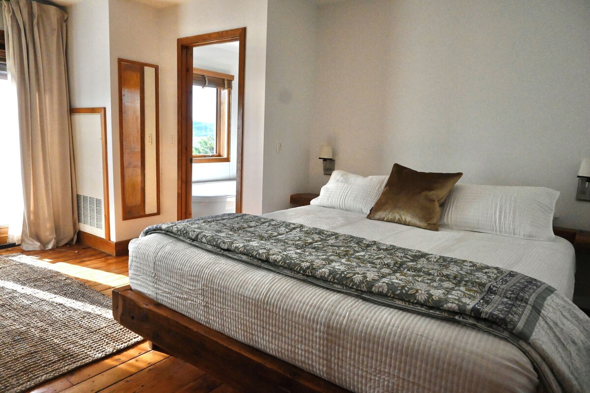Quarto com cama de madeira, colcha estampada, vista através de uma porta aberta e janela com cortinas.