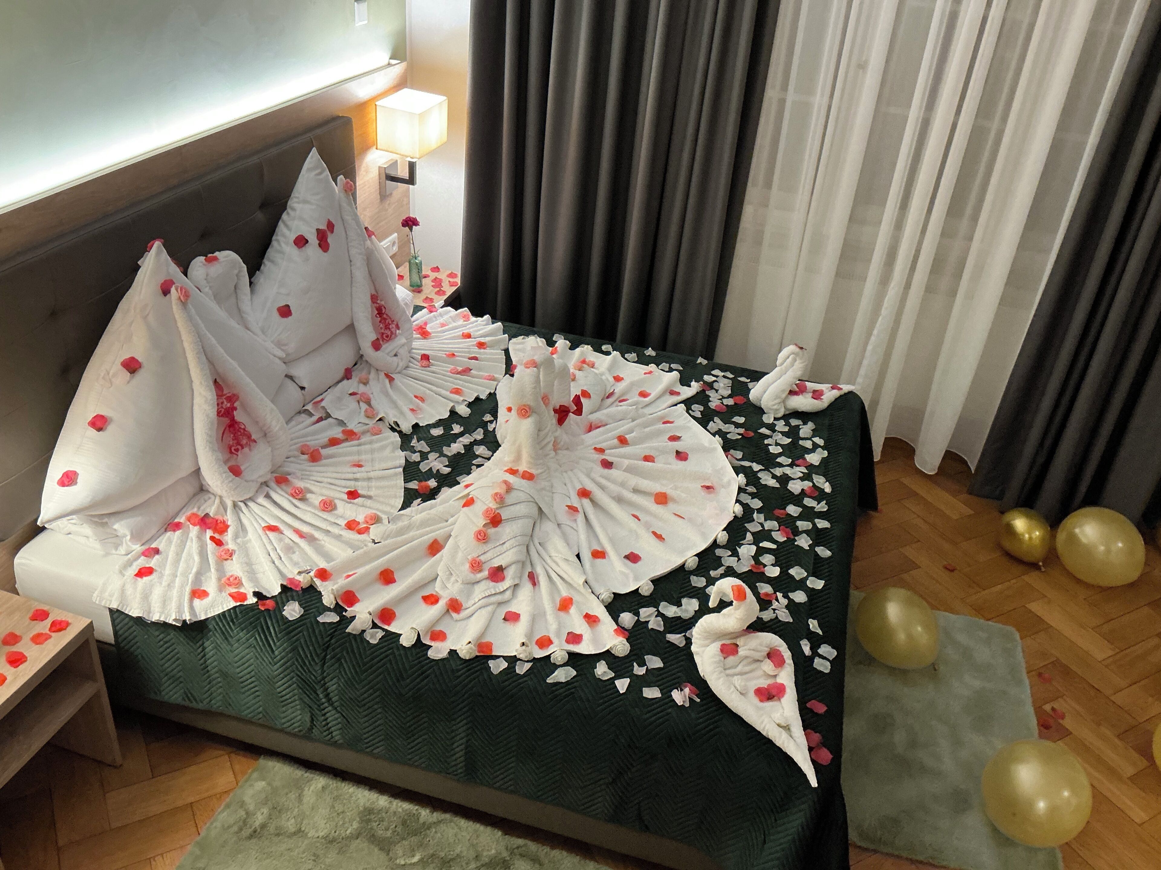 Cama com pétalas de rosa e decorações de cisne, mesa de cabeceira, luminária e vista do quarto.