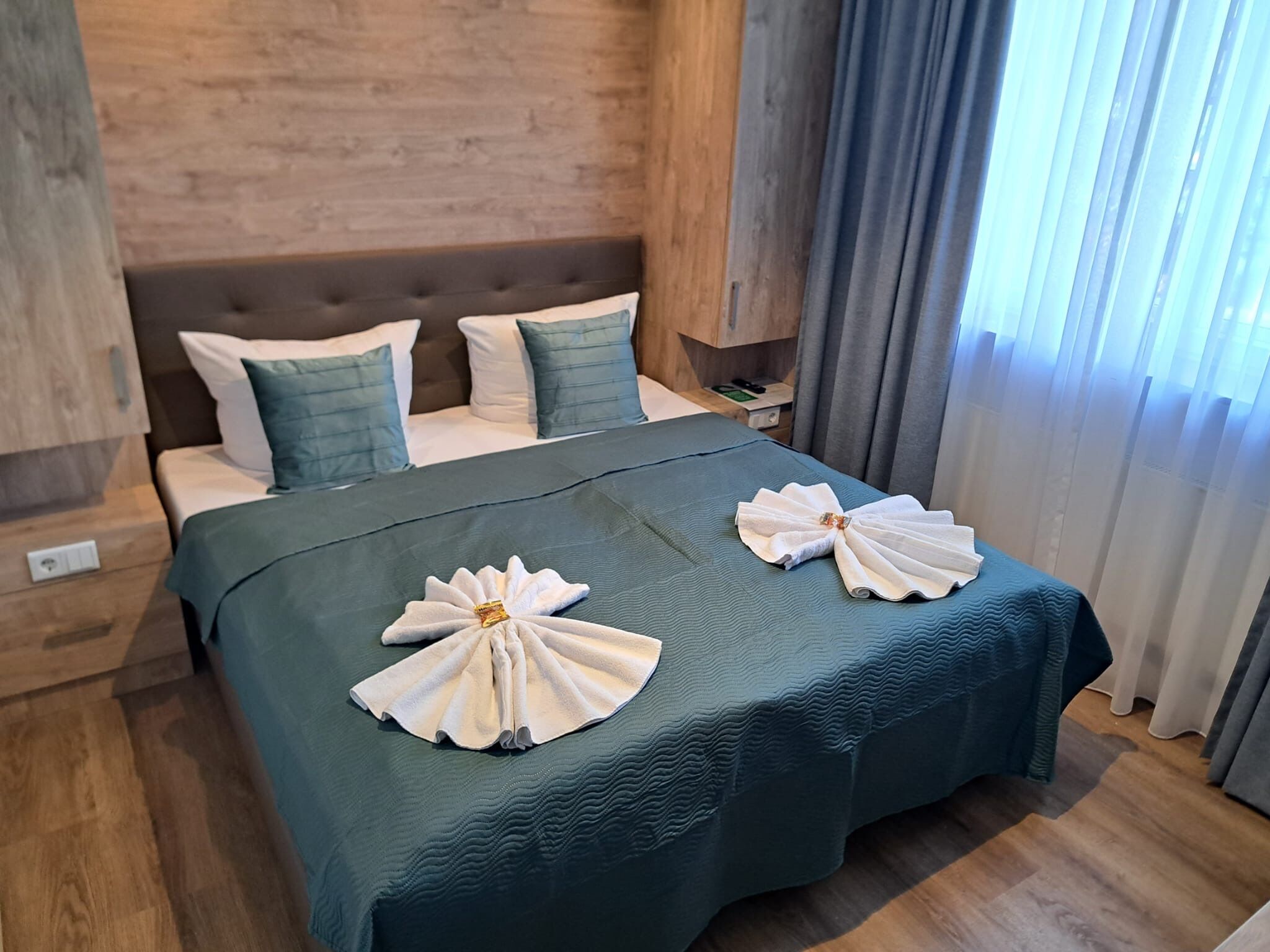 Quarto de hotel com uma cama, mesa de cabeceira, cabeceira de madeira e janela com cortinas.