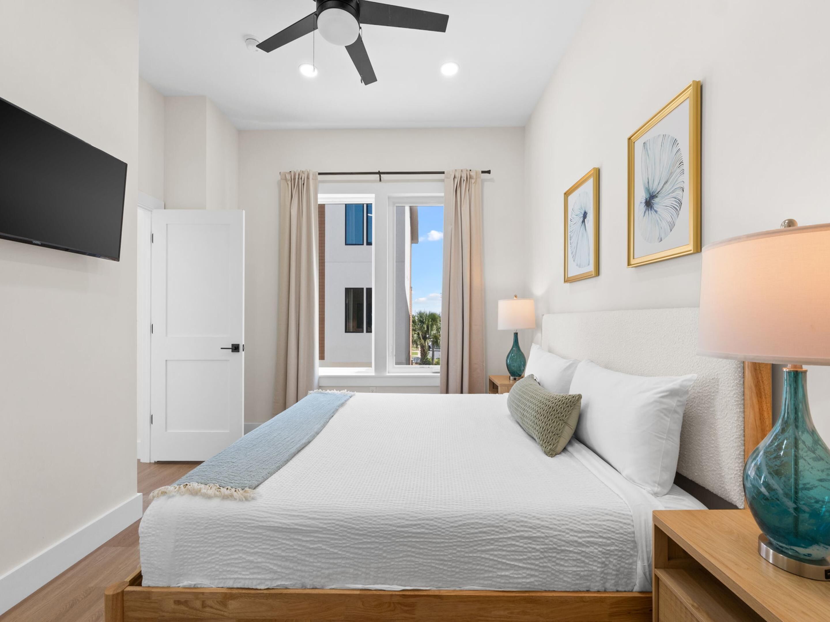 Quarto com cama, ventoinha no teto, televisão, janela com cortinas e parede com quadros emoldurados.