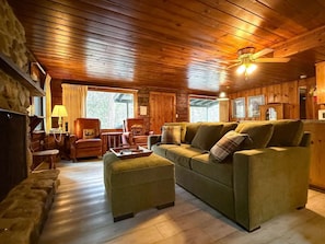 Interior acolhedor de uma cabana, com teto de madeira, um sofá, uma poltrona, uma mesa pequena e um abajur.