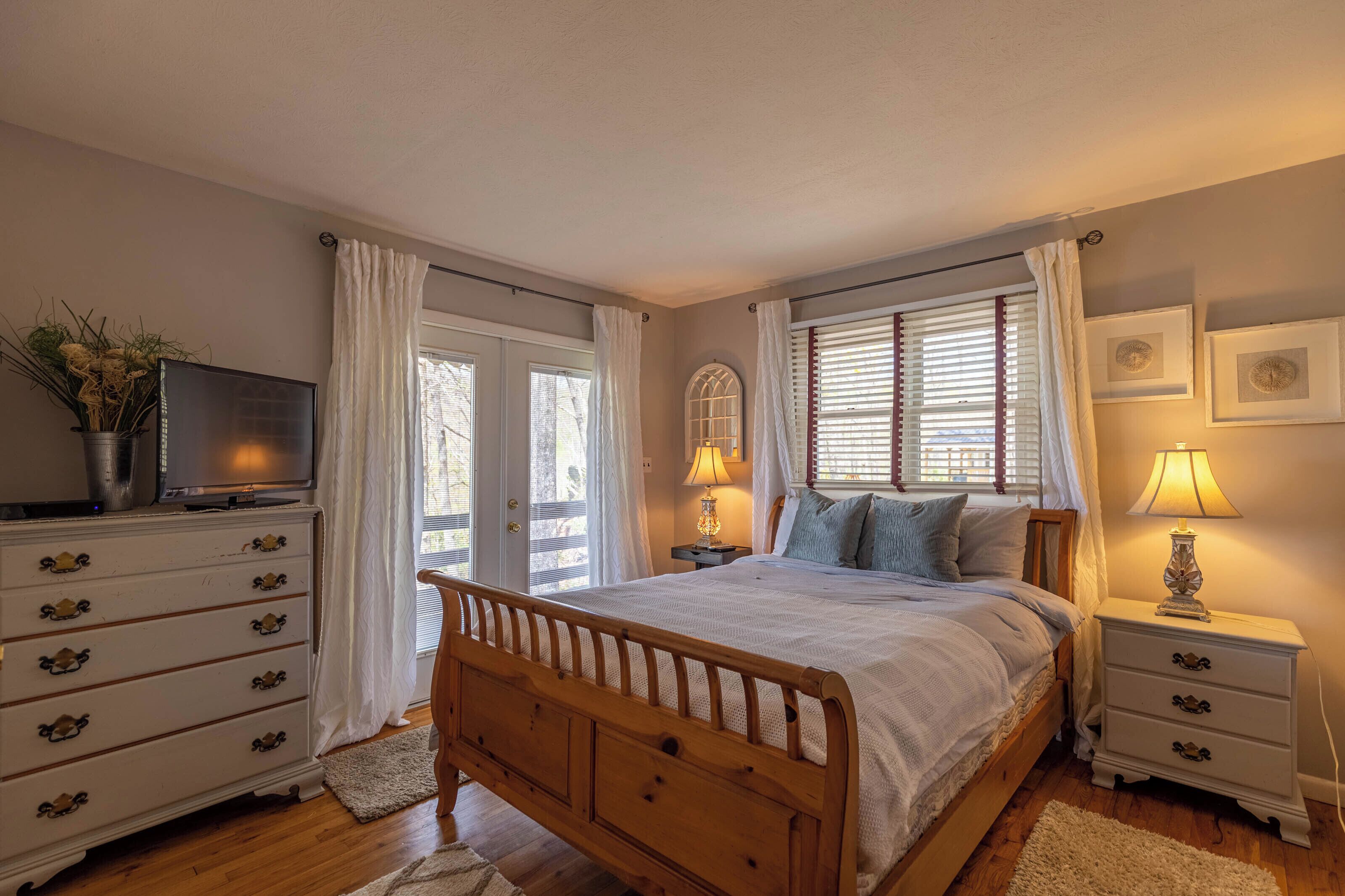 Quarto com cama de madeira, penteadeira, televisão e janela com cortinas.