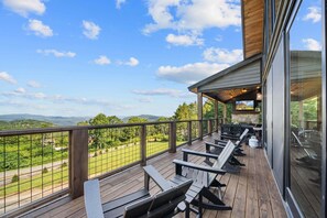 Um deck de madeira com área de estar externa, lareira de pedra e vista panorâmica para montanhas e vegetação.