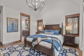 Quarto com uma cama grande, um candelabro, uma cabeceira de madeira, uma mesa de cabeceira, uma luminária e uma janela com persianas.