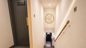 Um corredor com uma porta escura identificada com 'Apartment 2', uma escada de madeira com corrimão e um candeeiro geométrico na parede.
