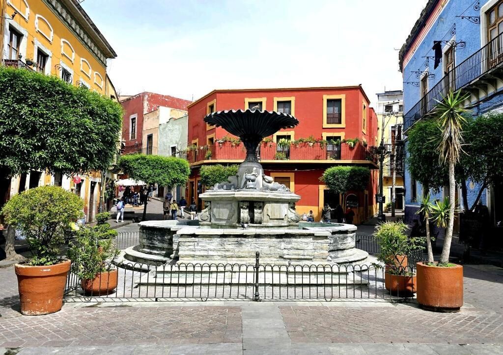 Uma fonte com uma estátua no centro, rodeada por um muro de pedra, numa praça com plantas em vasos e edifícios ao fundo.