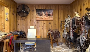 Um quarto com paredes de madeira, uma pintura de cavalos, um ventilador, uma mesa com cadeiras e equipamentos de equitação.