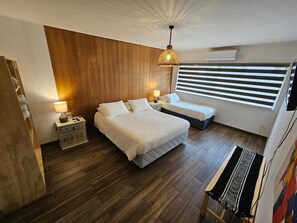 Um quarto com uma cama grande, uma cama menor, uma mesa de cabeceira, uma luminária, uma parede de madeira, uma janela com persianas e um lustre.