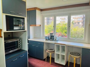 Uma cozinha compacta com micro-ondas, torradeira e cafeteira na bancada, uma janela com vista para área verde e um banquinho pequeno.