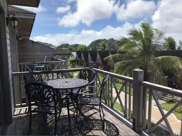 Um deck de madeira com mesa e cadeiras de metal, cercado por um corrimão, com vista para uma paisagem com palmeiras e o céu.