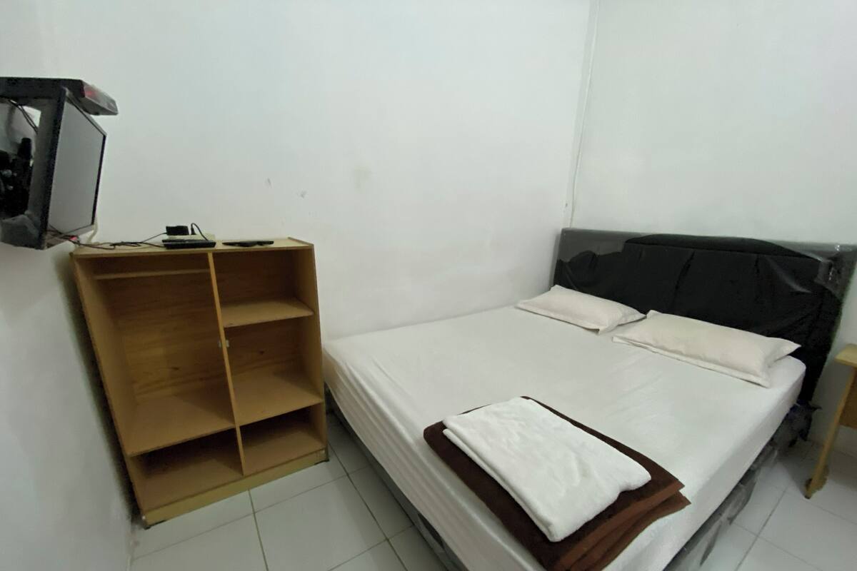 Uma cama de solteiro com roupa de cama branca, uma mesa de cabeceira de madeira e uma TV fixada na parede.