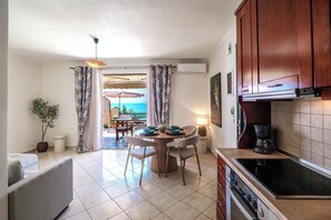 Uma cozinha moderna com armários de madeira, um espaço de refeições com mesa e cadeiras redondas, e vista para o mar através de uma porta de vidro.