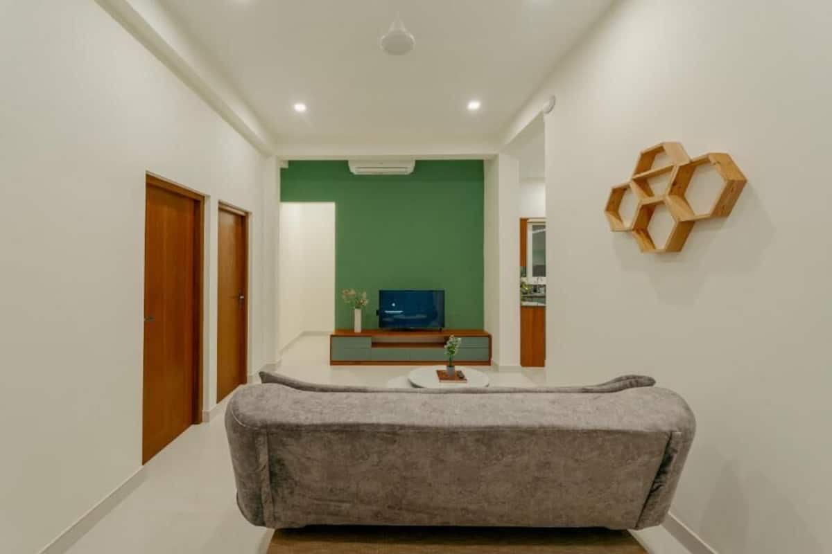 Um corredor com um sofá cinzento, uma televisão sobre um suporte de madeira e uma parede verde.