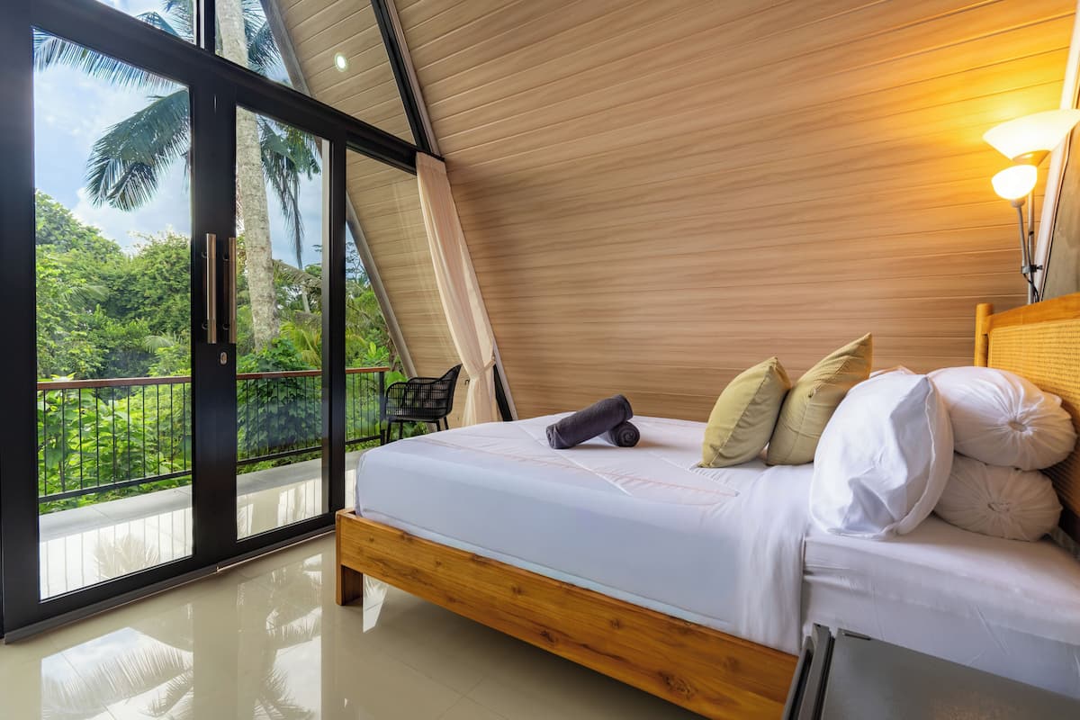 Quarto com cama, almofadas, mesa de cabeceira, luminária e vista para o verde através de uma porta de correr.