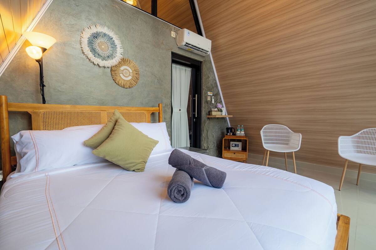 Uma cama bem arrumada com almofadas e toalhas, uma mesa de cabeceira com abajur, e um ar condicionado fixado na parede.
