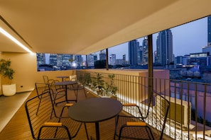 Varanda com vista para a cidade ao entardecer, incluindo uma mesa, cadeiras, um vaso de planta e um corrimão.