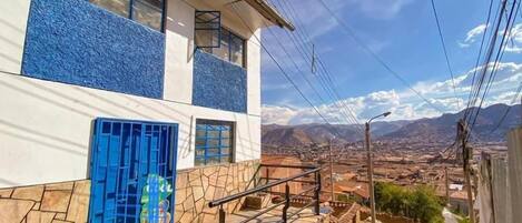 Um edifício com porta azul e janelas, uma parede de pedra e vista para um vale com montanhas ao fundo.