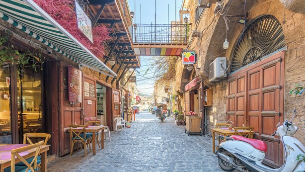 Uma rua de paralelepípedos com uma scooter estacionada, mesas e cadeiras de madeira e um edifício com uma placa.