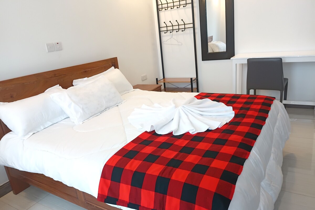 Cama bem arrumada com lençóis brancos, manta xadrez vermelha e preta e cabeceira de madeira.