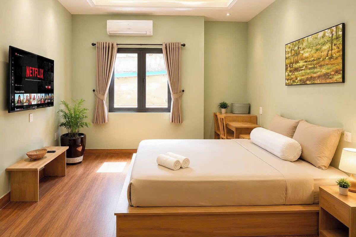 Quarto de hotel com cama, televisão exibindo Netflix, janela com cortinas, escrivaninha com uma planta e um quadro na parede.