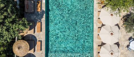 Vista aérea de uma piscina retangular com água cristalina, rodeada por espreguiçadeiras e guarda-sóis, adjacente a um edifício com telhado ladrilhado.
