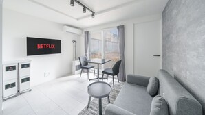 Uma sala de estar moderna com um sofá cinza, uma mesa redonda pequena, uma TV fixada na parede e uma área de jantar com cadeiras e mesa.