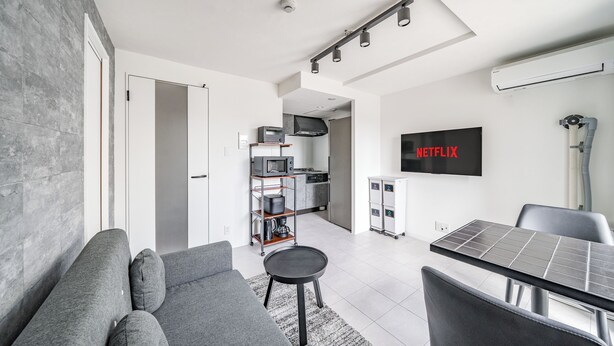 Uma sala moderna com um sofá cinzento, uma mesa de jantar com cadeiras, uma cozinha com eletrodomésticos e uma TV fixada na parede.