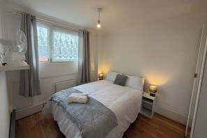 Quarto com cama, luminárias de cabeceira, janela com cortinas, radiador e ventoinha fixada na parede.