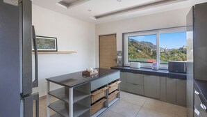 Uma cozinha moderna com geladeira, mesa de vidro, cestos de vime e vista para uma paisagem verde através de uma janela.
