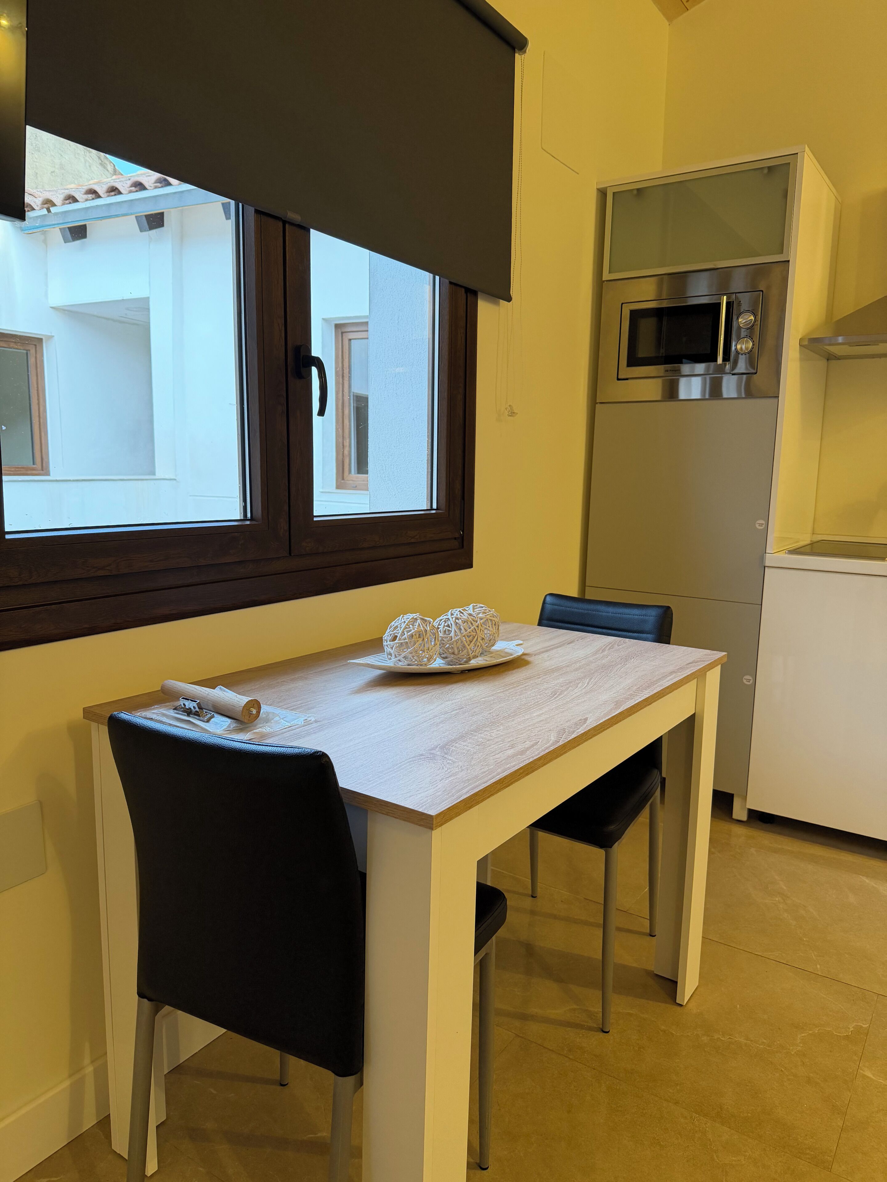 Uma cozinha pequena com micro-ondas, uma mesa com cadeiras e uma janela com vista para prédios.