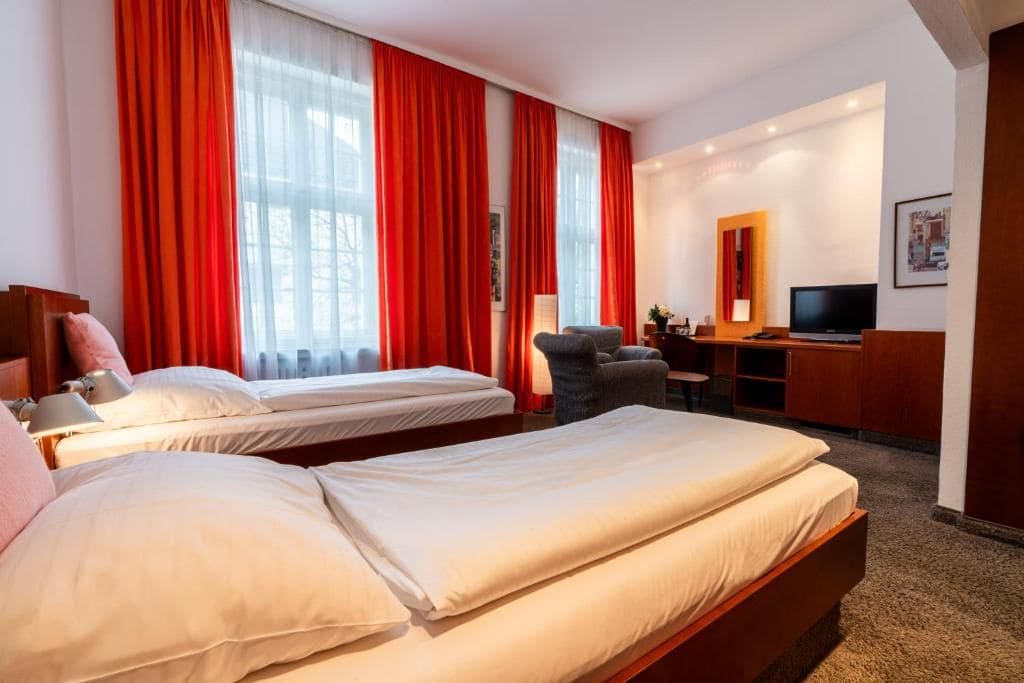 Quarto de hotel com duas camas, uma escrivaninha, uma cadeira, televisão e uma janela grande com cortinas vermelhas.